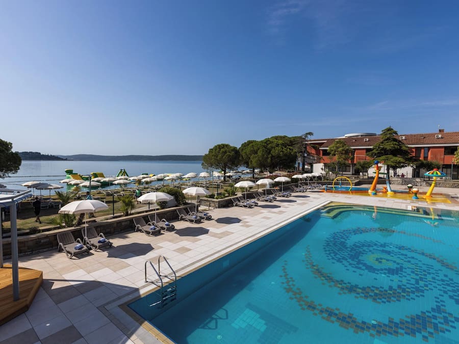 Uma área de piscina com espreguiçadeiras e guarda-sóis, vista clara do mar e um edifício com telhado ladrilhado.