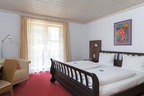 Quarto de hotel com cama, poltrona, escrivaninha, abajur e janela com cortinas.