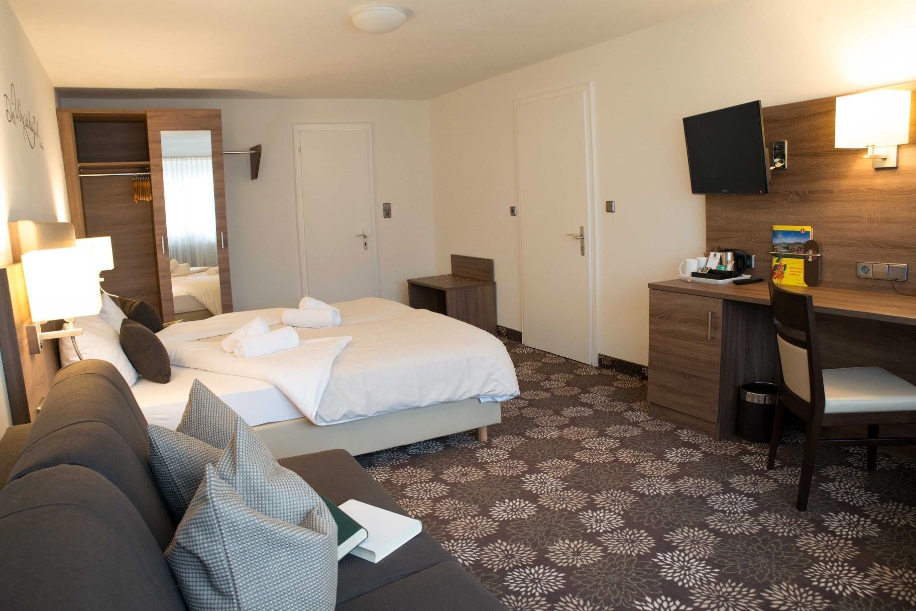 Quarto de hotel com cama, escrivaninha, cadeira, sofá e televisão.