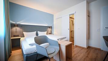 Quarto de hotel moderno com cama, escrivaninha, cadeira e área de chuveiro.