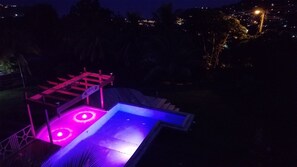 Piscina com deck de madeira e pérgola, rodeada por uma paisagem escura com luzes ao longe.