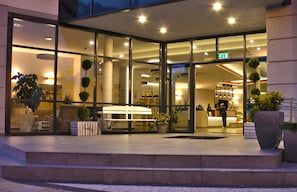Entrada de hotel com portas de vidro, balcão de recepção e plantas em vasos.