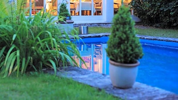 Piscina com uma planta em vaso no primeiro plano e um edifício com área de estar ao ar livre ao fundo.