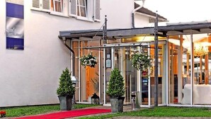 Entrada de hotel com tapete vermelho, cestos de flores pendentes e plantas em vasos.
