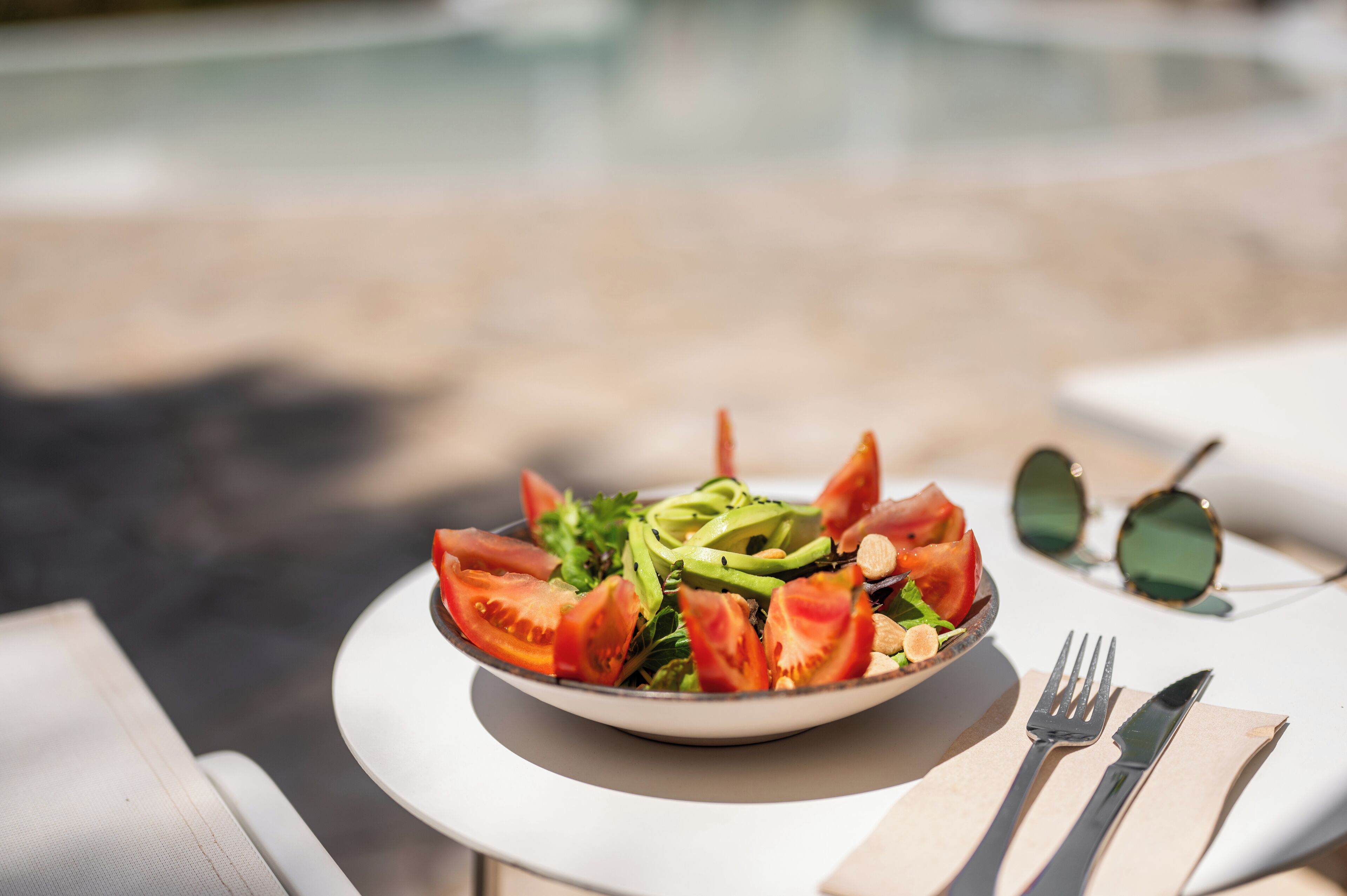 Uma salada num prato, com garfo e faca, sobre uma mesa perto de uma piscina.