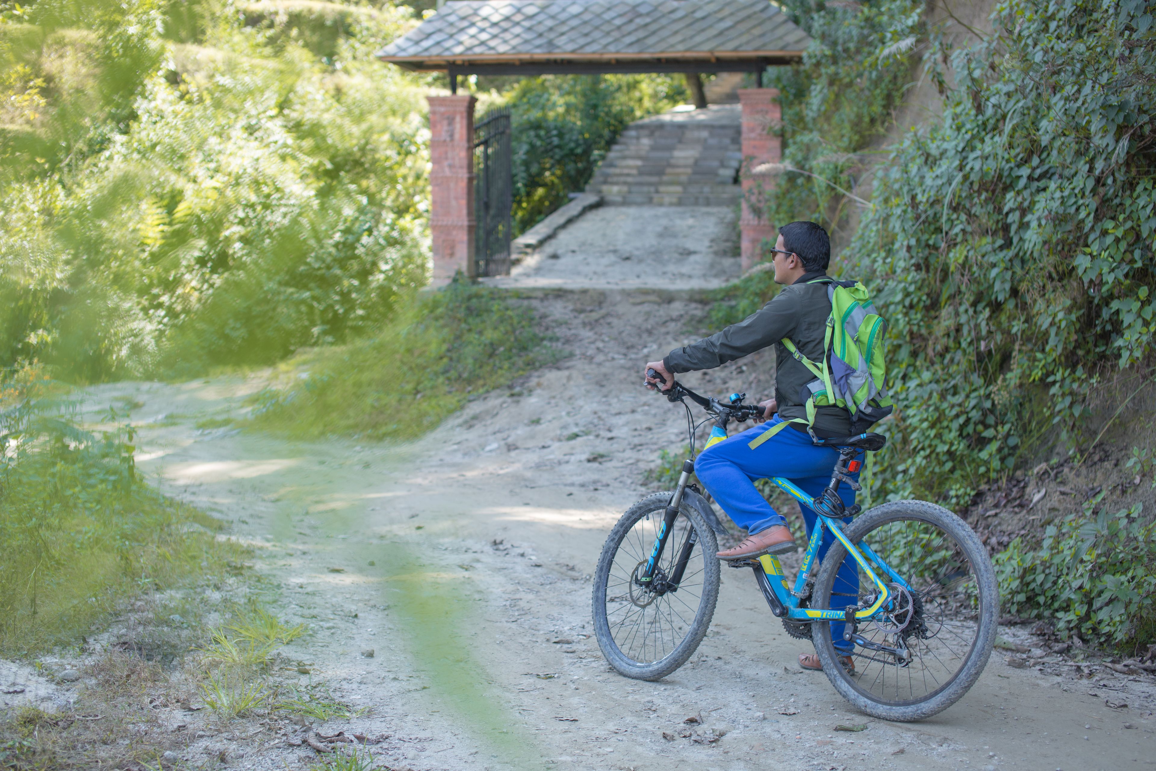 Uma pessoa anda de bicicleta de montanha por uma trilha de terra, com uma construção de tijolos ao fundo.