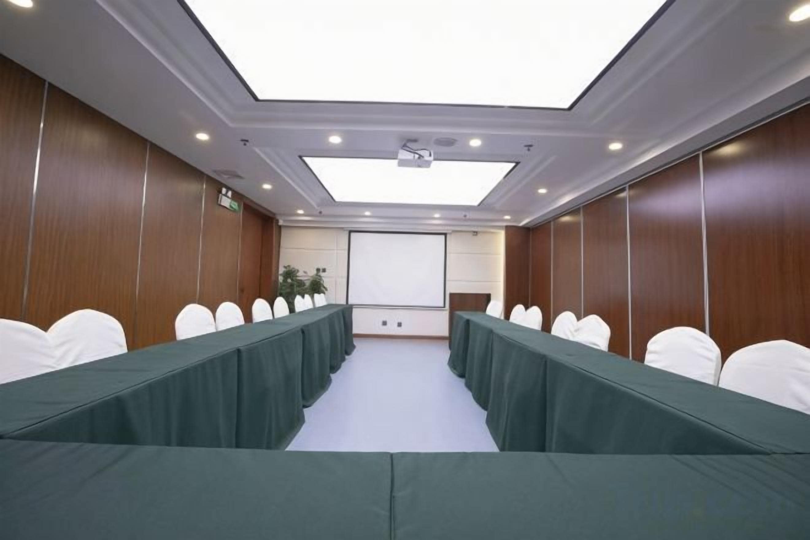Uma sala de conferências com uma mesa comprida, cadeiras brancas, uma tela de projetor e um claraboia.