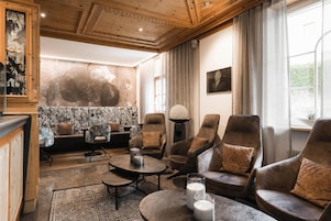 Quarto com teto de madeira, parede estampada e uma janela grande com cortinas. Há várias poltronas e um sofá.