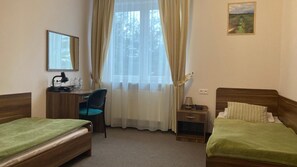Quarto com duas camas, uma escrivaninha, uma cadeira, janela com cortinas, um quadro na parede e uma mesinha de cabeceira com abajur.