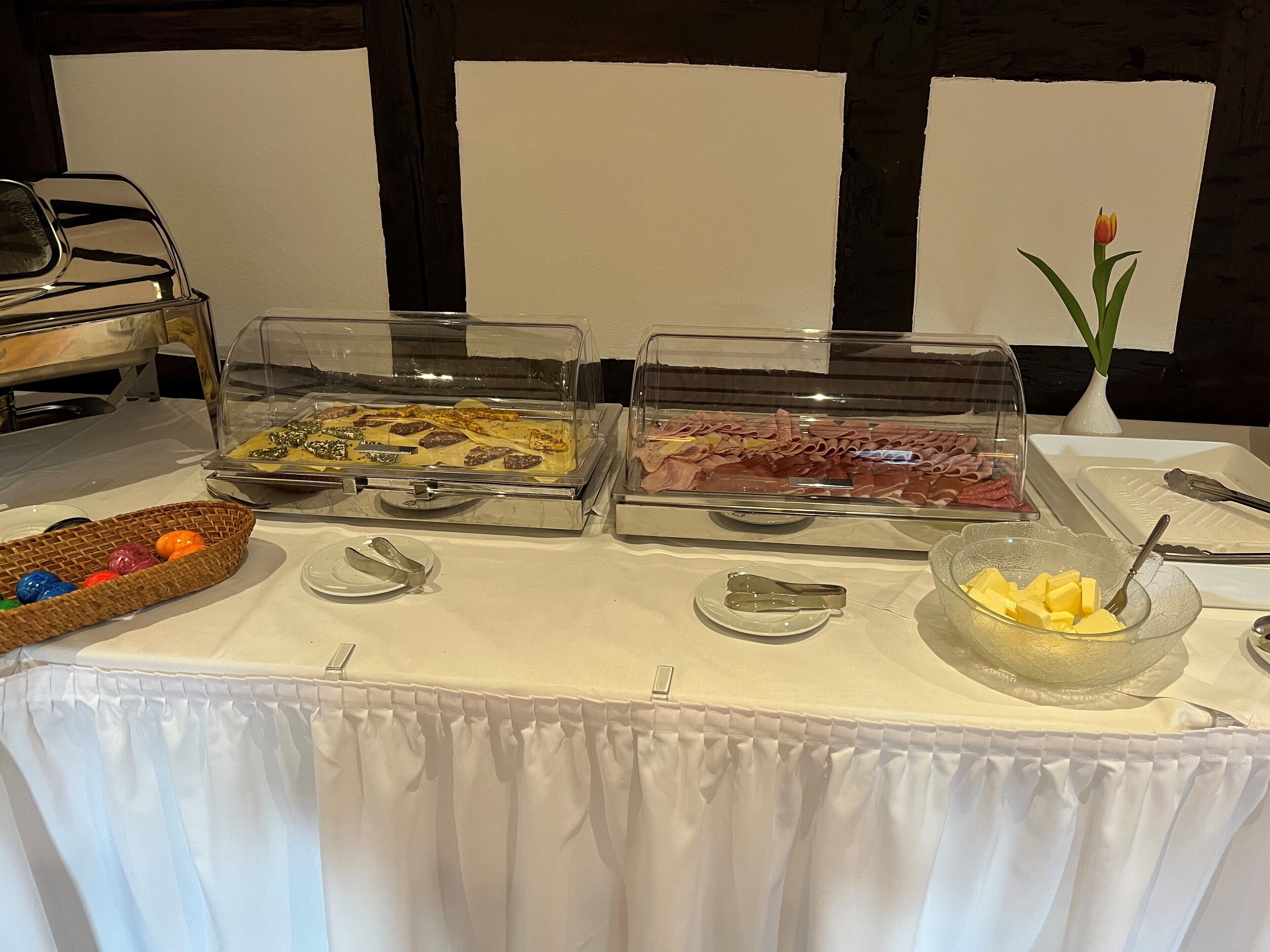 Uma mesa de buffet com diversos alimentos, incluindo uma cesta de ovos coloridos, uma tigela de manteiga e duas vitrines com alimentos.