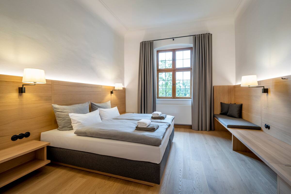 Quarto de hotel moderno com uma cama grande, mesas de cabeceira, um banco e uma janela com cortinas.