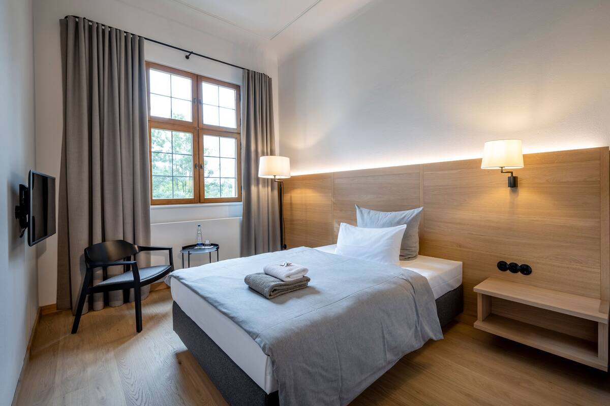 Quarto de hotel com uma cama, uma escrivaninha, uma cadeira, janela com cortinas e duas luminárias na parede.
