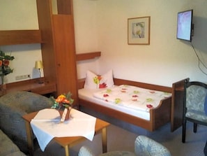 Quarto pequeno e acolhedor com uma cama de solteiro, uma cadeira, uma mesa com um vaso de flores e uma televisão na parede.