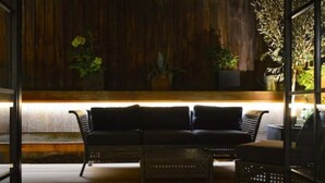 Uma sala de estar moderna com sofá escuro, cadeiras de vime, piso de madeira e parede de madeira com plantas em vasos.