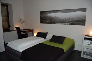 Quarto de hotel com cama, escrivaninha, cadeira, mesa de cabeceira e quadro na parede.