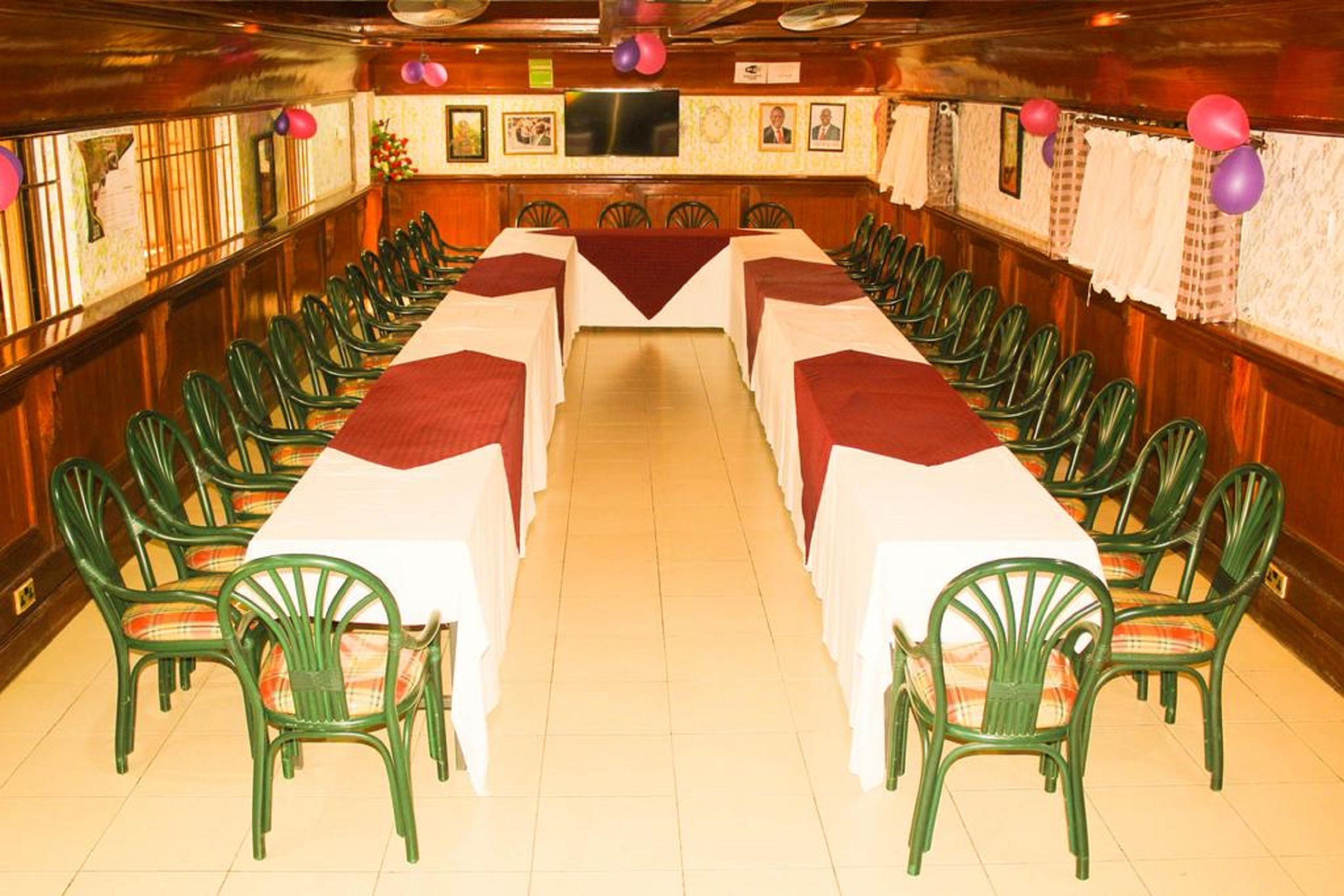 Uma longa sala de banquetes com fileiras de cadeiras verdes e mesas brancas, decorada com balões e quadros nas paredes.