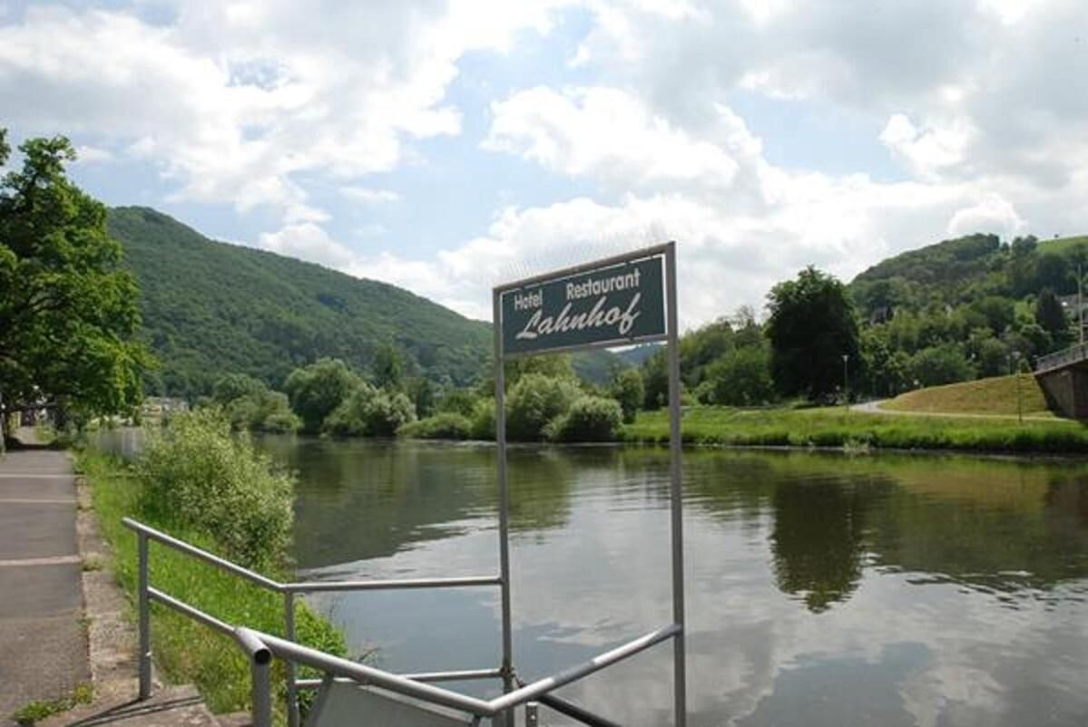 Placa de um hotel e restaurante chamado 'Lahnhof', próximo a um rio calmo, com guarda-corpo de metal e um caminho de acesso.