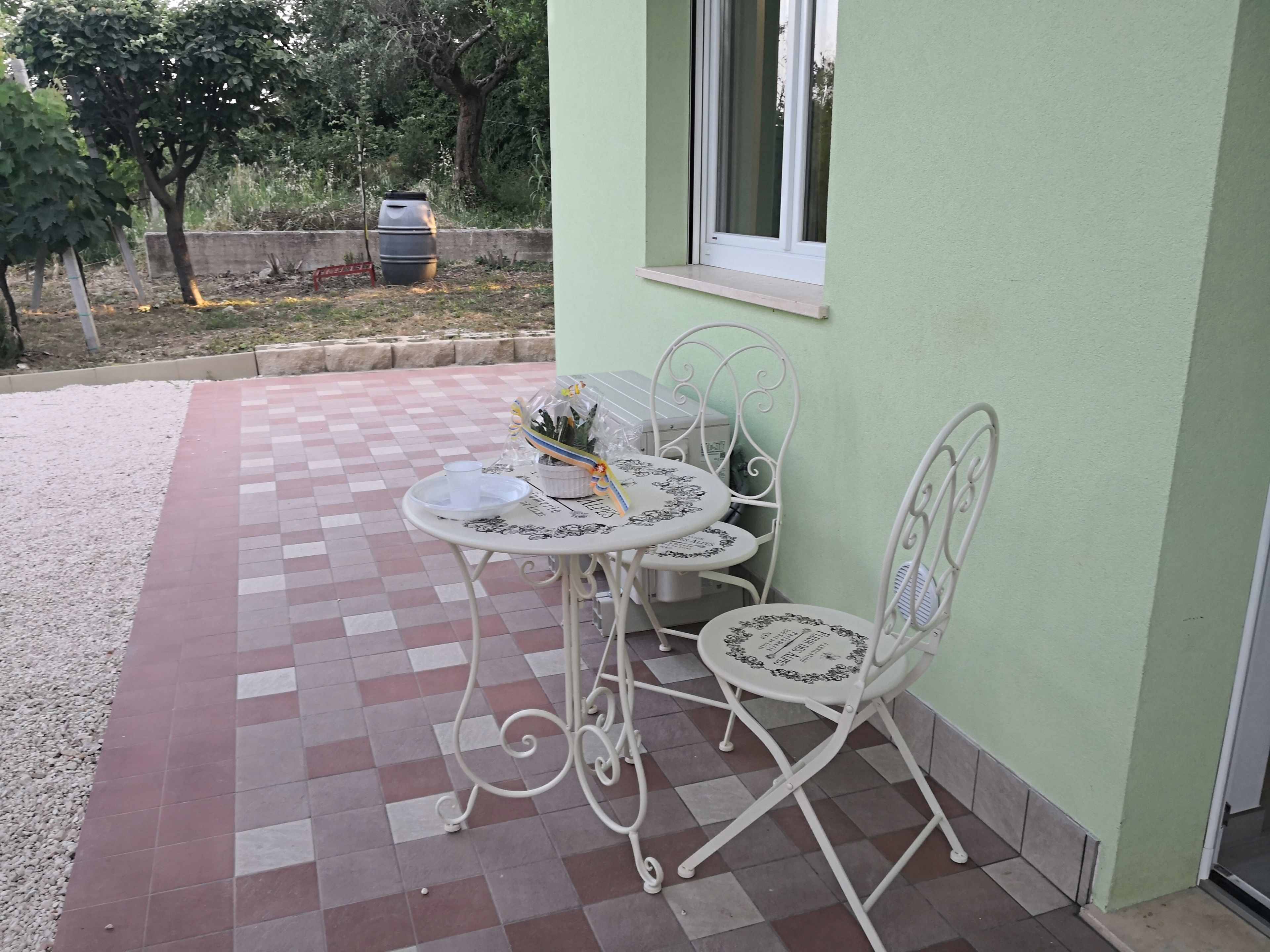 Um pátio com piso xadrez, uma mesa de metal branca com cadeiras e um pequeno vaso de planta.