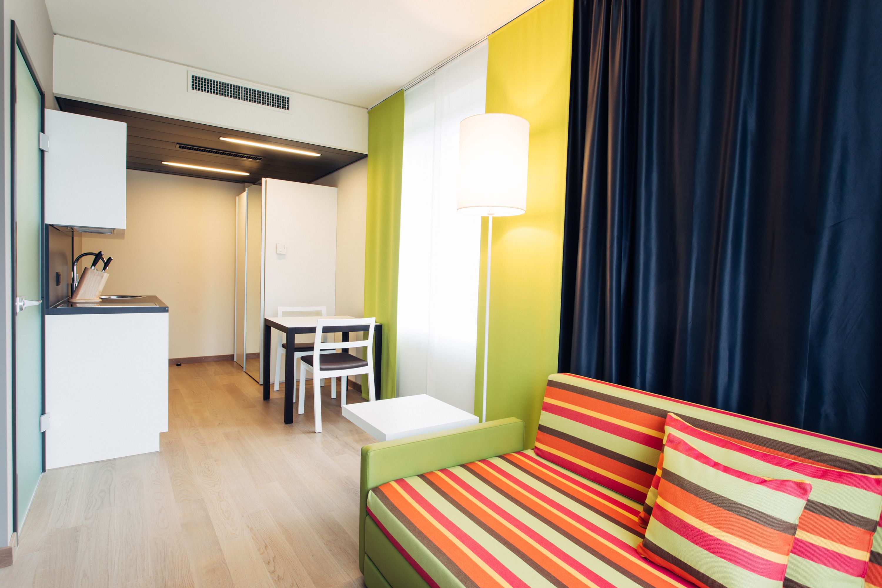 Quarto de hotel moderno com sofá verde, uma pequena mesa branca, uma escrivaninha e uma luminária.