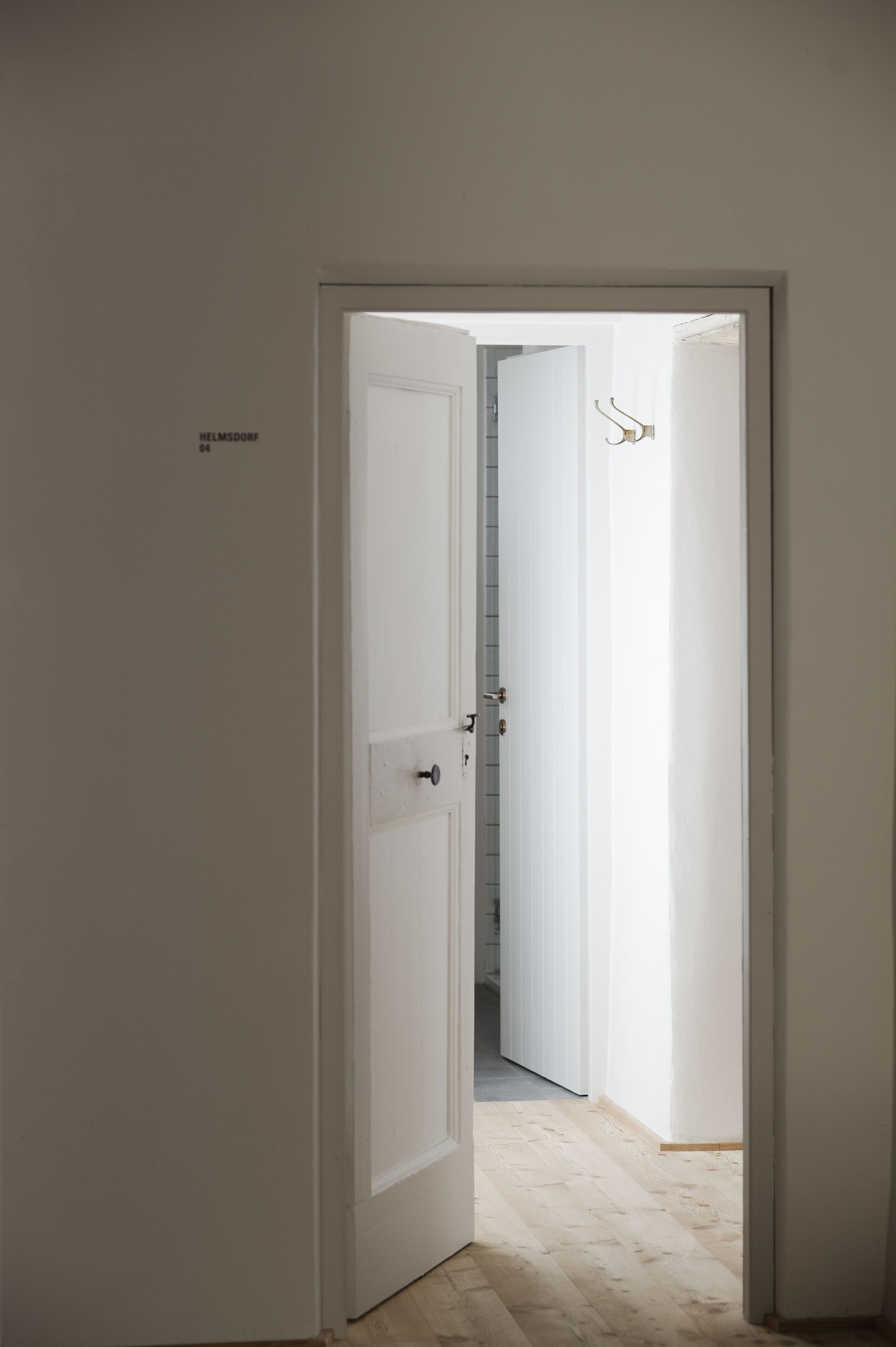 Uma porta branca com uma pequena placa acima, dando acesso a um corredor com soalho de madeira.