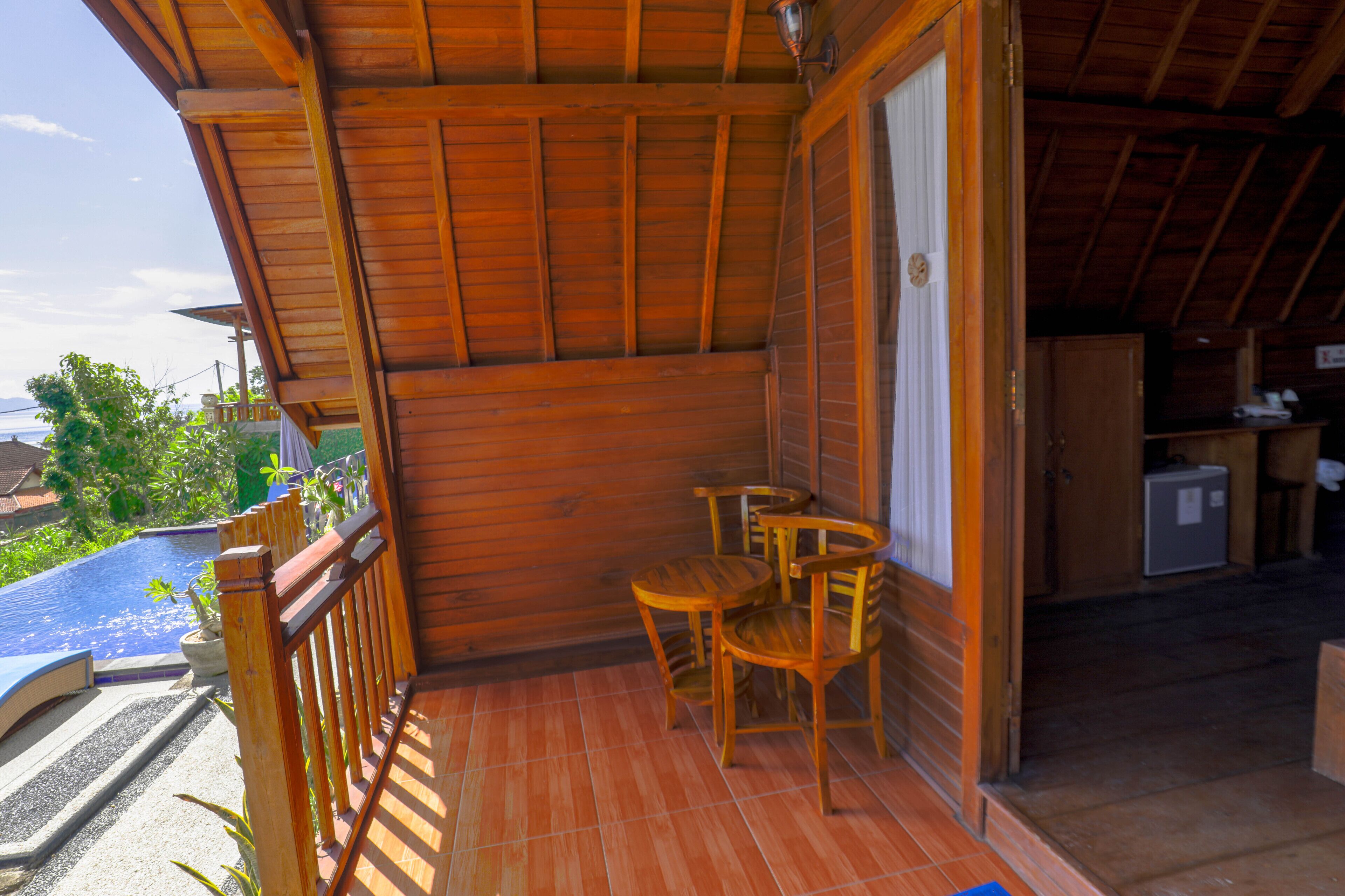 Varanda de madeira com vista para uma piscina, mobiliário de madeira e porta que dá para um quarto.