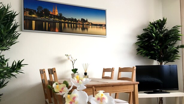 Uma sala de jantar com uma mesa e cadeiras de madeira, uma televisão sobre um suporte e um quadro grande na parede.