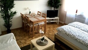 Quarto de hotel com uma mesa de jantar de madeira, cadeiras, uma TV em um suporte, uma cama com roupa de cama estampada e duas plantas em vasos.