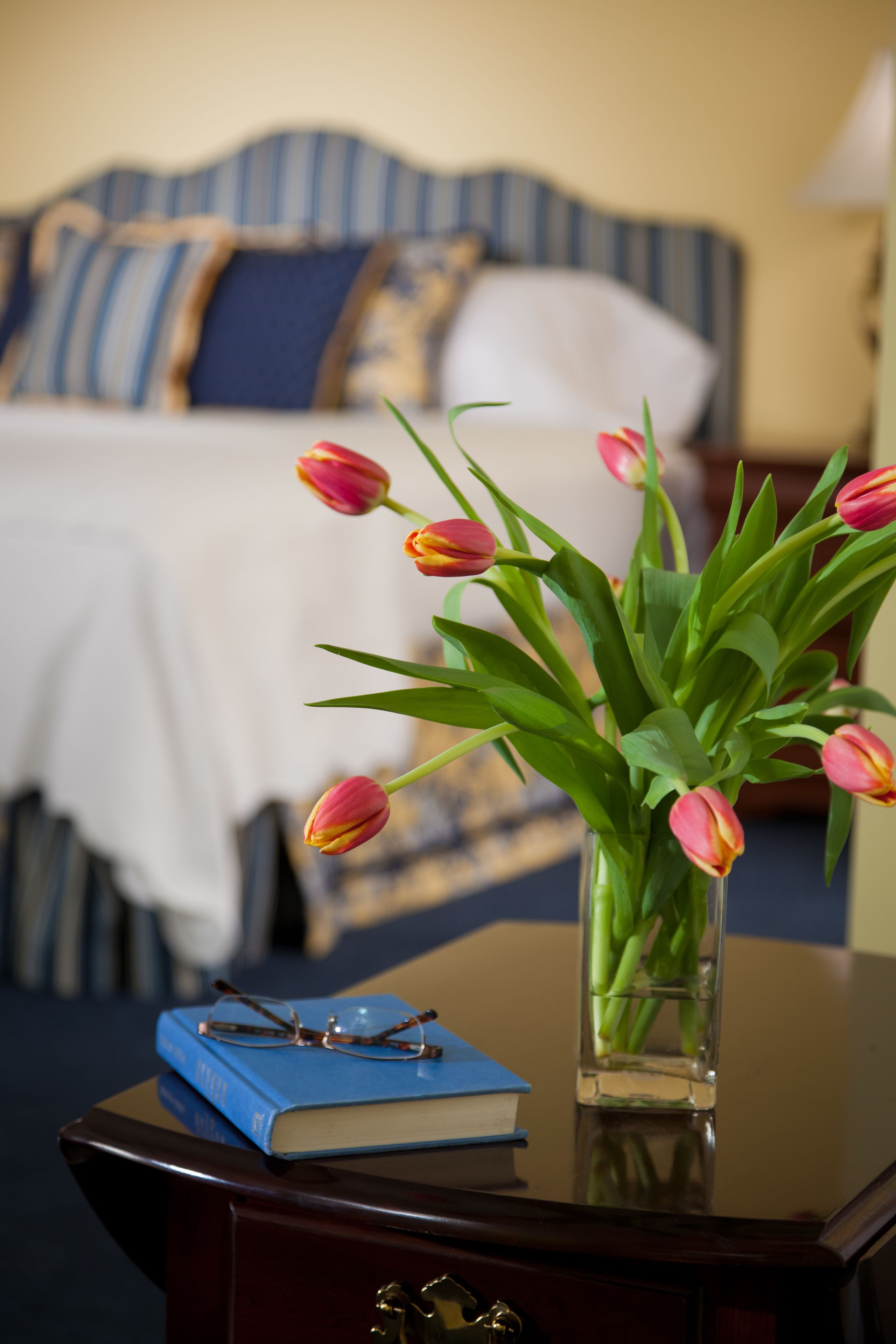 Uma cama com roupa de cama e almofadas listradas, uma mesa de cabeceira com um livro e óculos, e um vaso com tulipas.