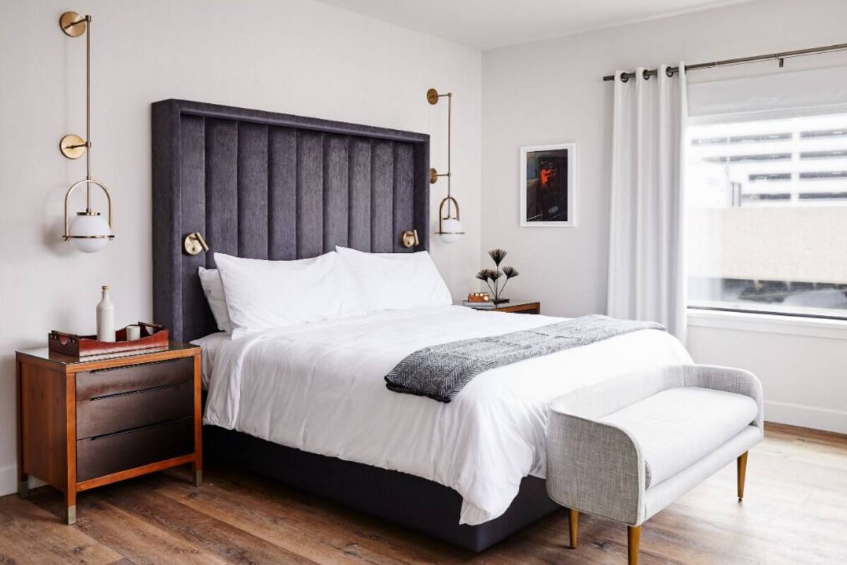 Quarto moderno com cabeceira escura, roupa de cama branca, mesa de cabeceira com abajur e vista para o exterior pela janela.