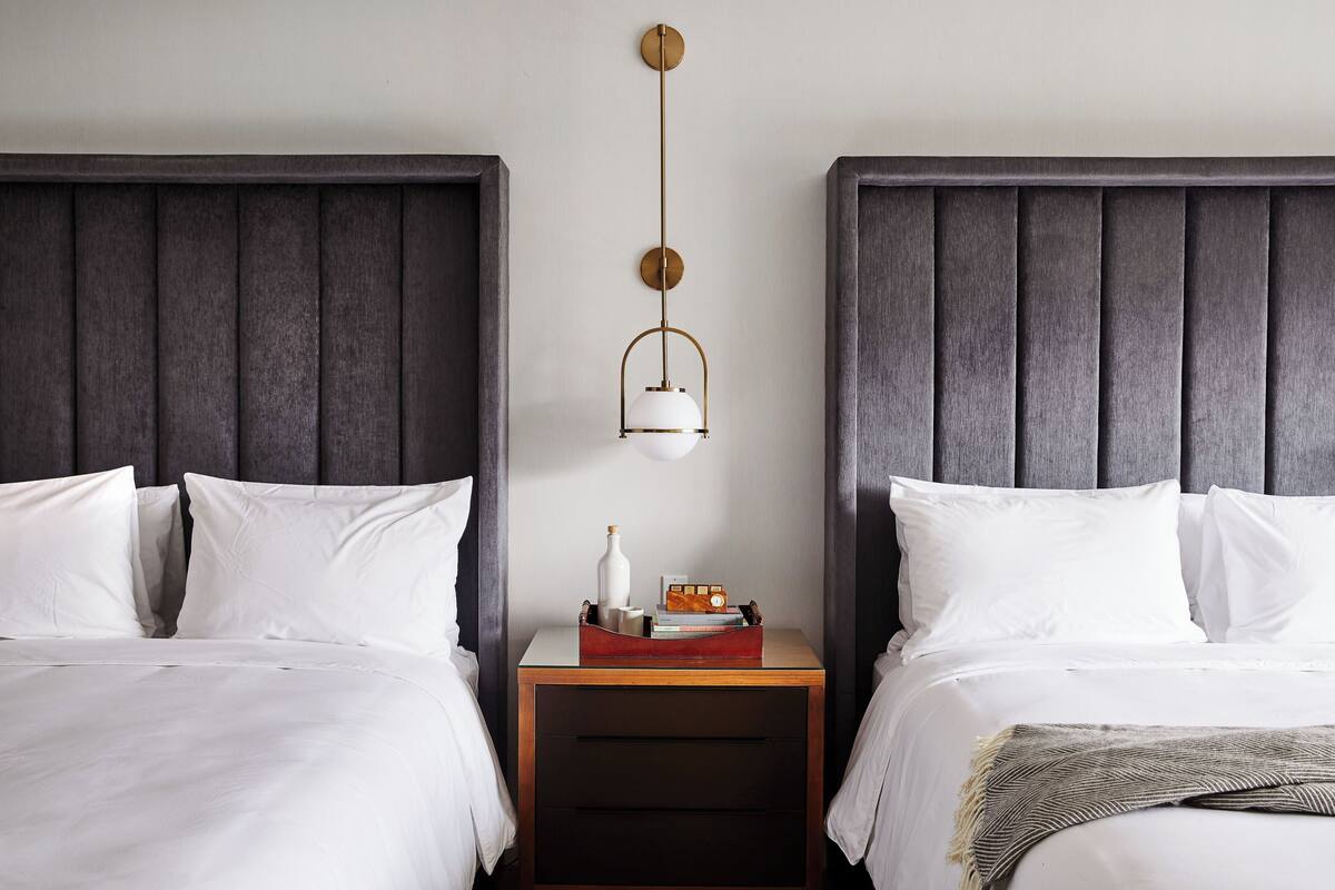Quarto de hotel com duas camas, uma mesa de cabeceira de madeira, um abajur branco e uma mesinha com um livro e uma garrafa.