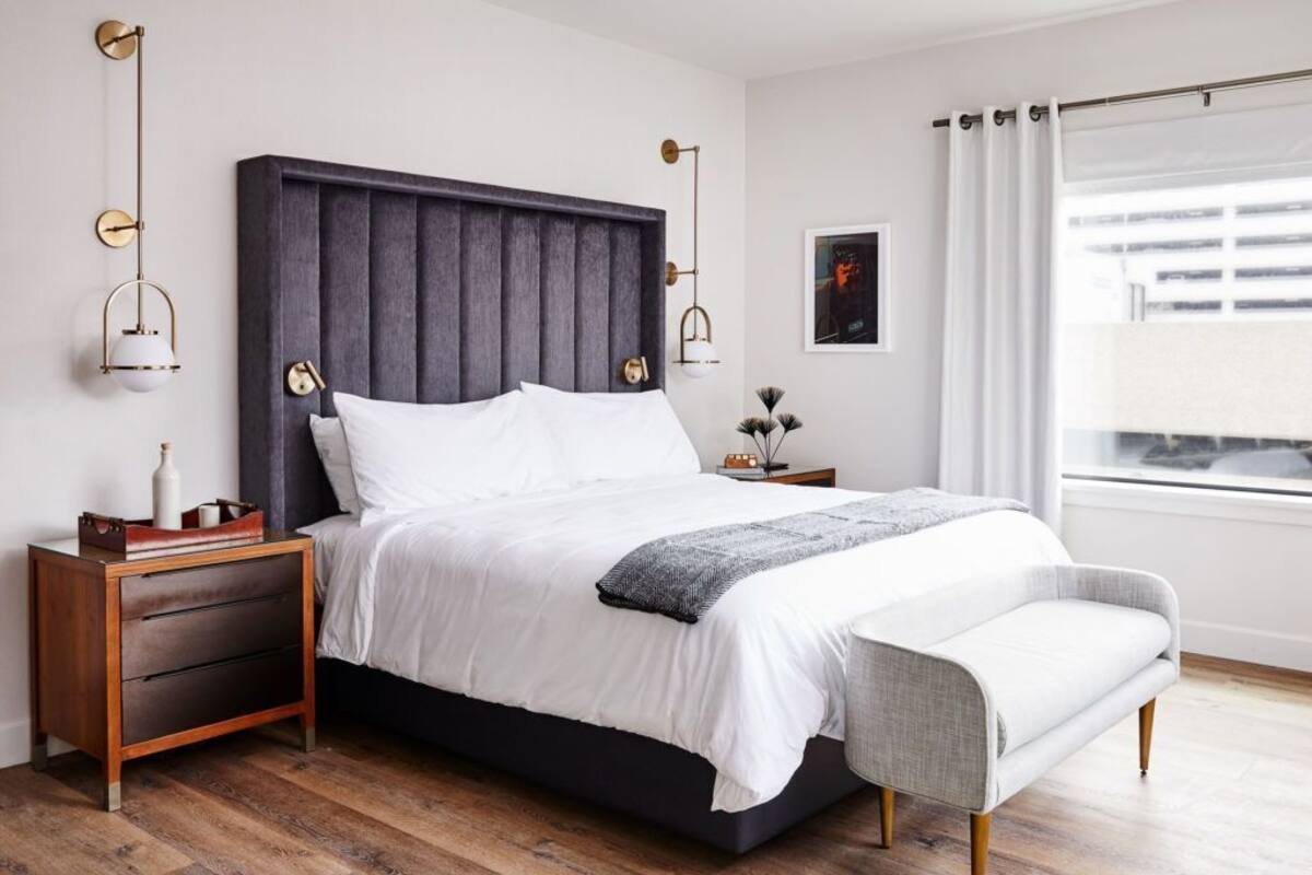 Quarto moderno com cabeceira escura, roupa de cama branca, mesa de cabeceira com gavetas, luminária e janela com cortinas.