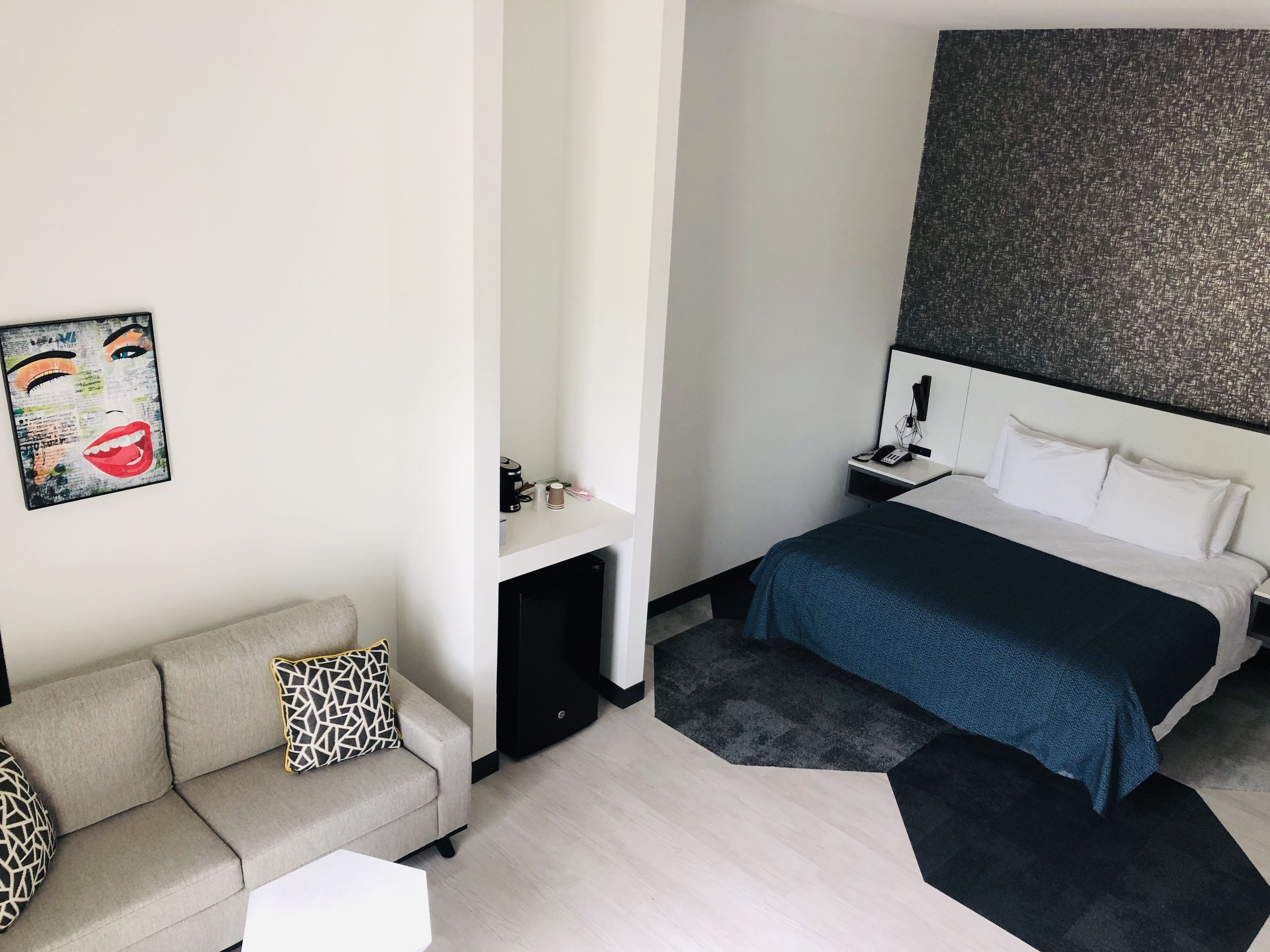 Quarto de hotel moderno com cama, sofá, mesinha e televisão fixada na parede.