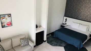 Quarto de hotel moderno com cama, sofá, mesinha e televisão fixada na parede.