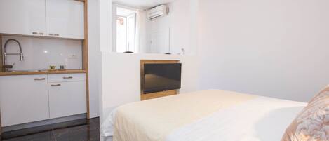 Quarto de hotel moderno com uma cama, uma televisão fixada na parede, uma pequena área de cozinha com pia e armários, e um espelho sobre a pia.
