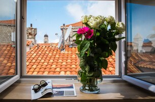 Uma janela com vista para telhados de telha de barro, um edifício branco com cúpula e um vaso com flores no parapeito.