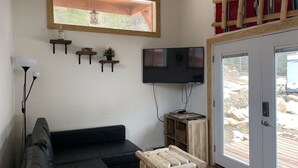Uma sala de estar com sofá preto, mesa de centro de madeira, TV fixada na parede e uma porta de vidro deslizante que dá para o exterior.