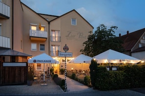 Um hotel chamado 'Zur Post' com mesas ao ar livre e guarda-sóis, uma placa indicando 'Biergarten' e uma tenda da cervejaria Paulaner.