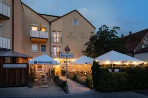 Um hotel chamado 'Zur Post' com mesas ao ar livre e guarda-sóis, uma placa indicando 'Biergarten' e uma tenda da cervejaria Paulaner.