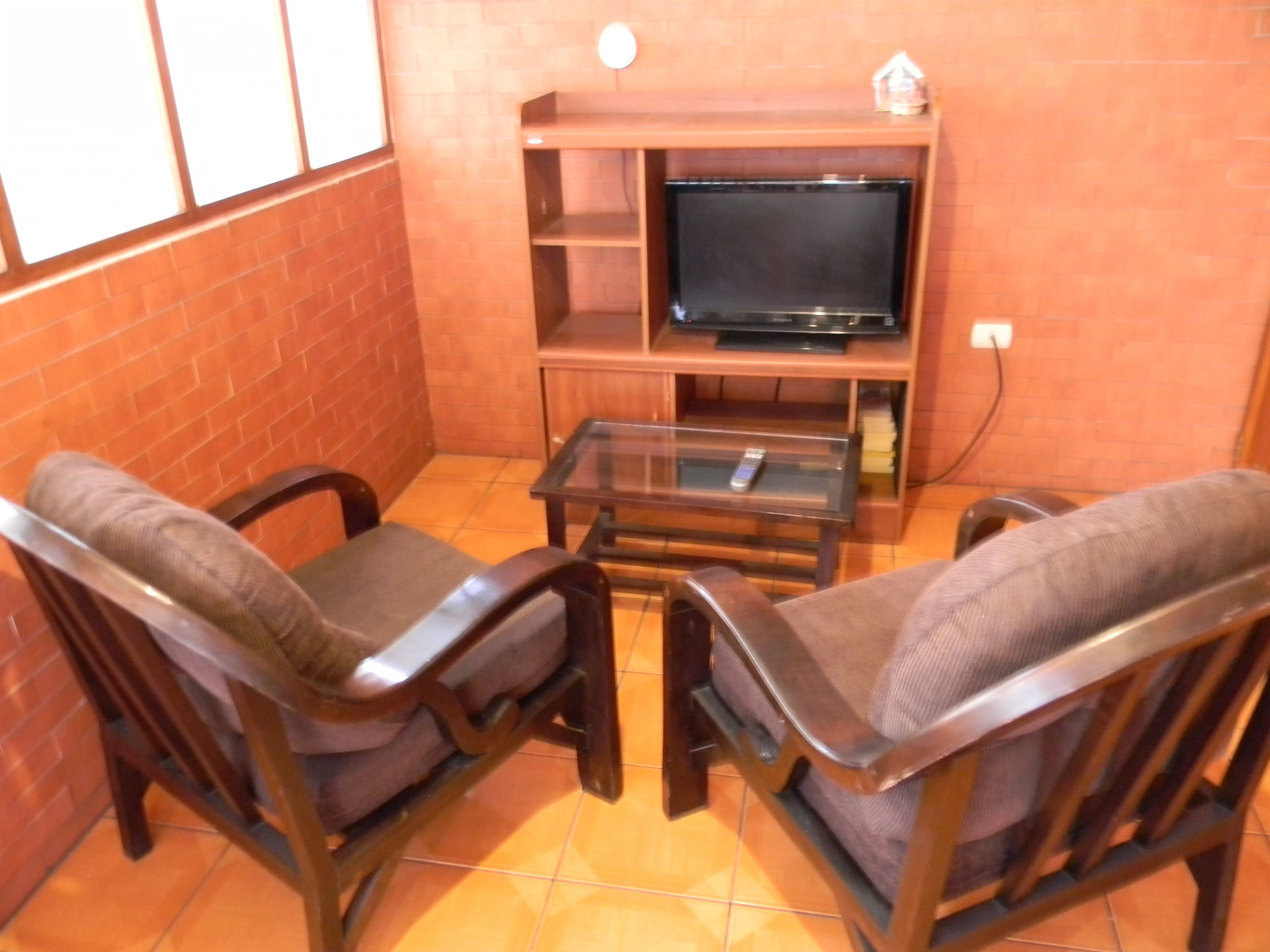 Uma sala de estar com duas cadeiras de madeira, uma mesa de centro de vidro e uma TV sobre um móvel de madeira.