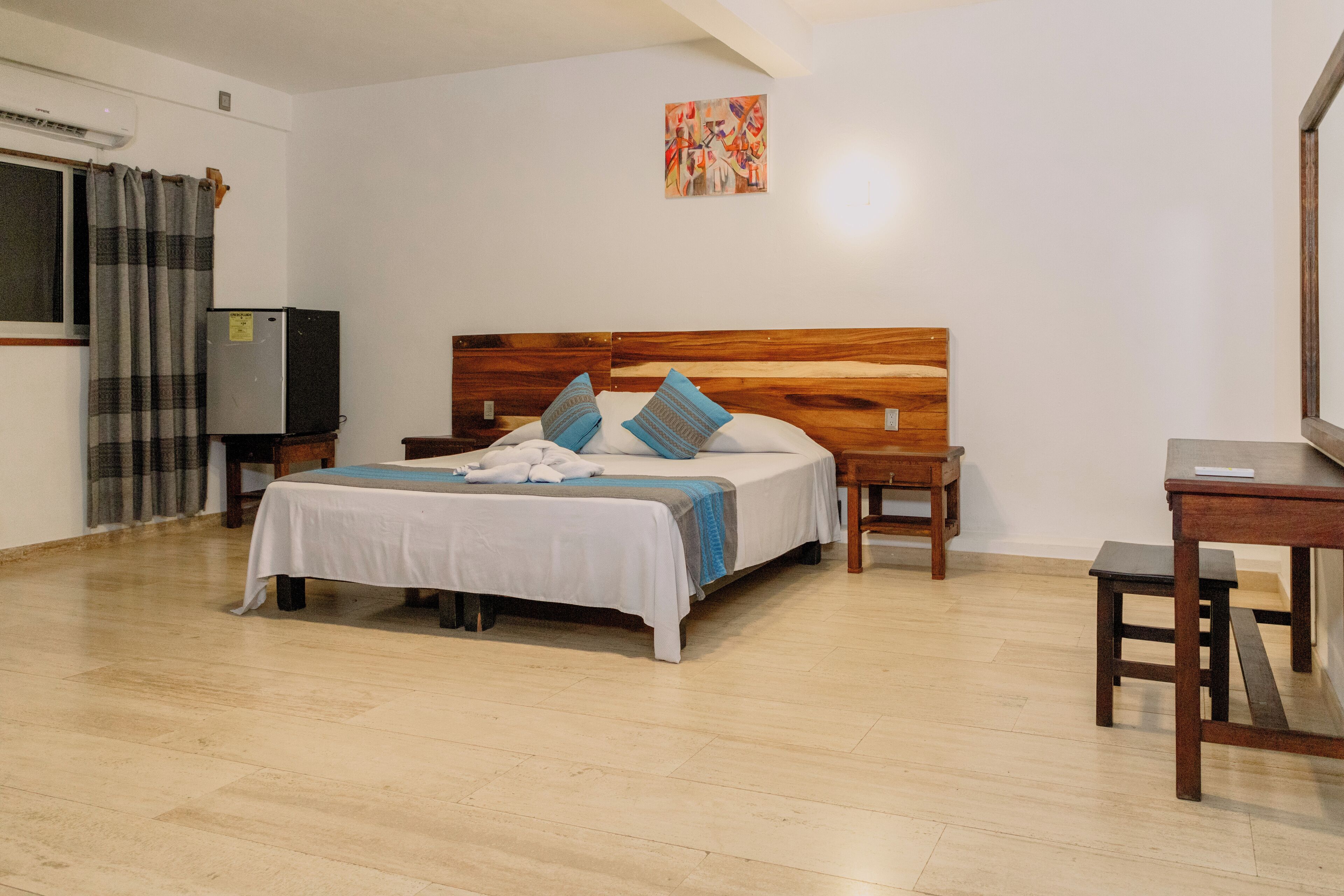 Quarto de hotel com cama de madeira, uma mesinha, uma cadeira, televisão e frigorífico.