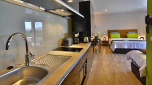 Quarto de hotel moderno com cozinha, uma cama e almofadas verdes, e um telefone ao lado da cama.