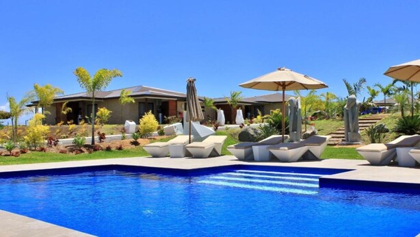 Piscina com espreguiçadeiras e guarda-sóis, rodeada por jardins ajardinados e um edifício com telhado inclinado.