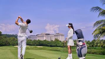 Um campo de golfe com um jogador se preparando para dar a tacada, um carregador com uma mala de golfe e um hotel ao fundo.