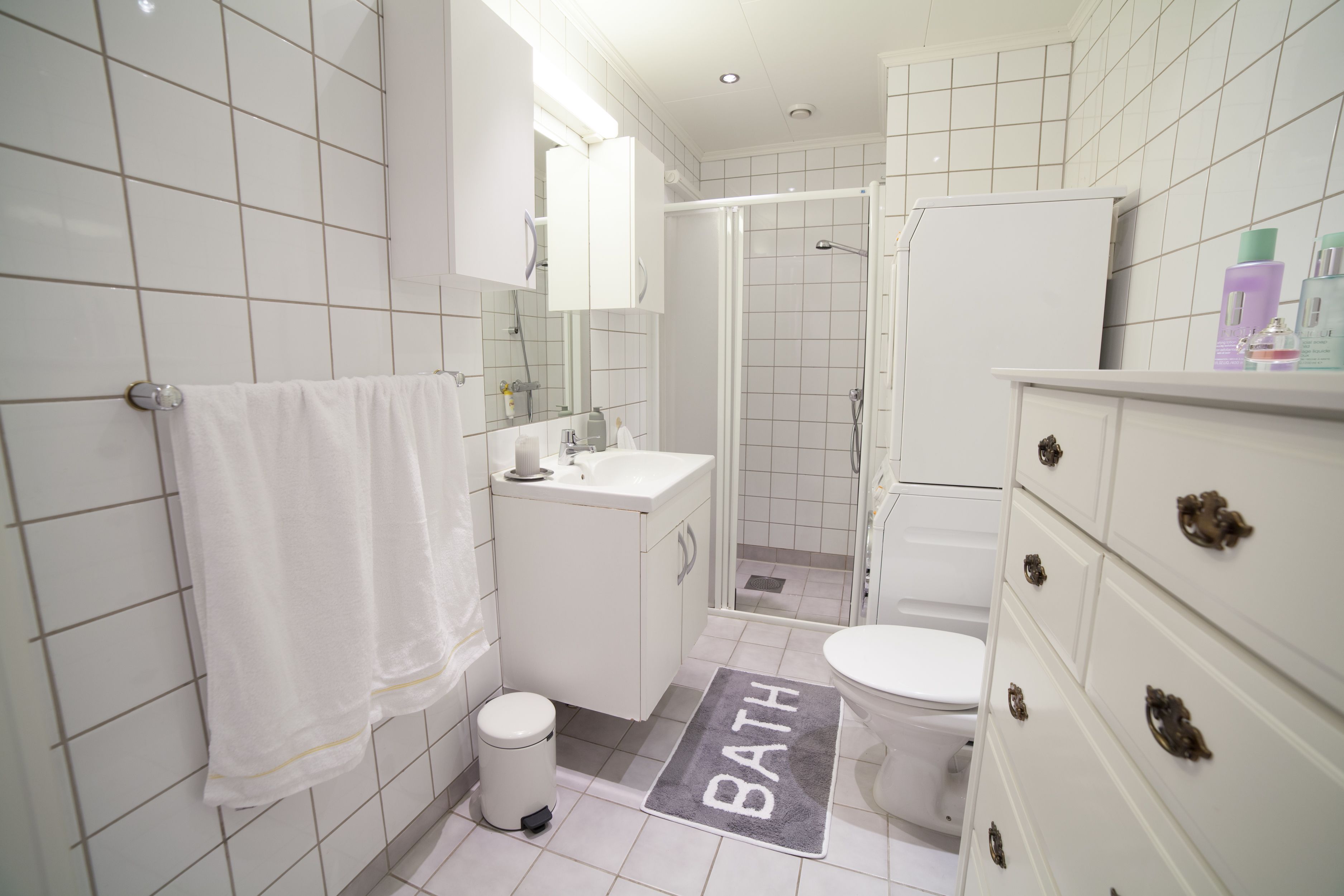 Casa de banho moderna com azulejos brancos, lavatório, sanita, chuveiro e toucador com gavetas.