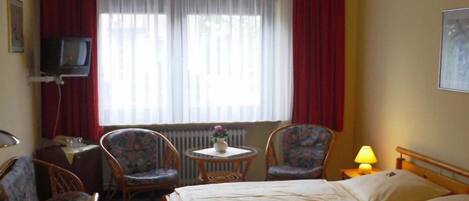Quarto de hotel com uma cama, duas cadeiras, uma mesinha com um vaso, uma luminária e uma televisão sobre um suporte.