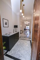 Um corredor com piso de mármore, um balcão preto com luminária e um quadro na parede.