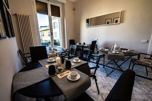 Uma sala de jantar com uma mesa posta para quatro pessoas, incluindo pratos, copos e talheres. Cadeiras cercam a mesa, e uma janela oferece vista para o exterior.