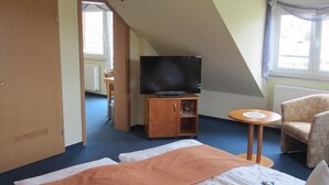 Quarto de hotel com cama, televisão sobre um móvel de madeira, uma mesinha redonda e uma cadeira.
