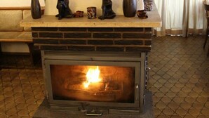 Lareira de pedra com fogo aceso, uma estante acima com objetos decorativos, e um chão com padrão.