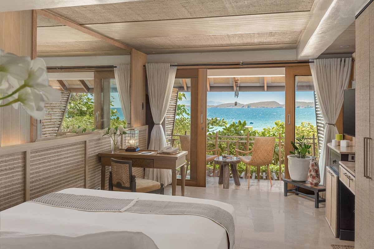 Quarto de hotel moderno com uma cama grande, uma mesa com abajur, uma pequena mesa com cadeiras e vista para o mar.